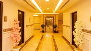Um hall de hotel com piso de mármore, duas plantas em vasos e uma escada que leva a um elevador.