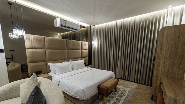 Quarto de hotel moderno com uma cama grande, cabeceira, um banco, uma mesinha e um candeeiro suspenso.