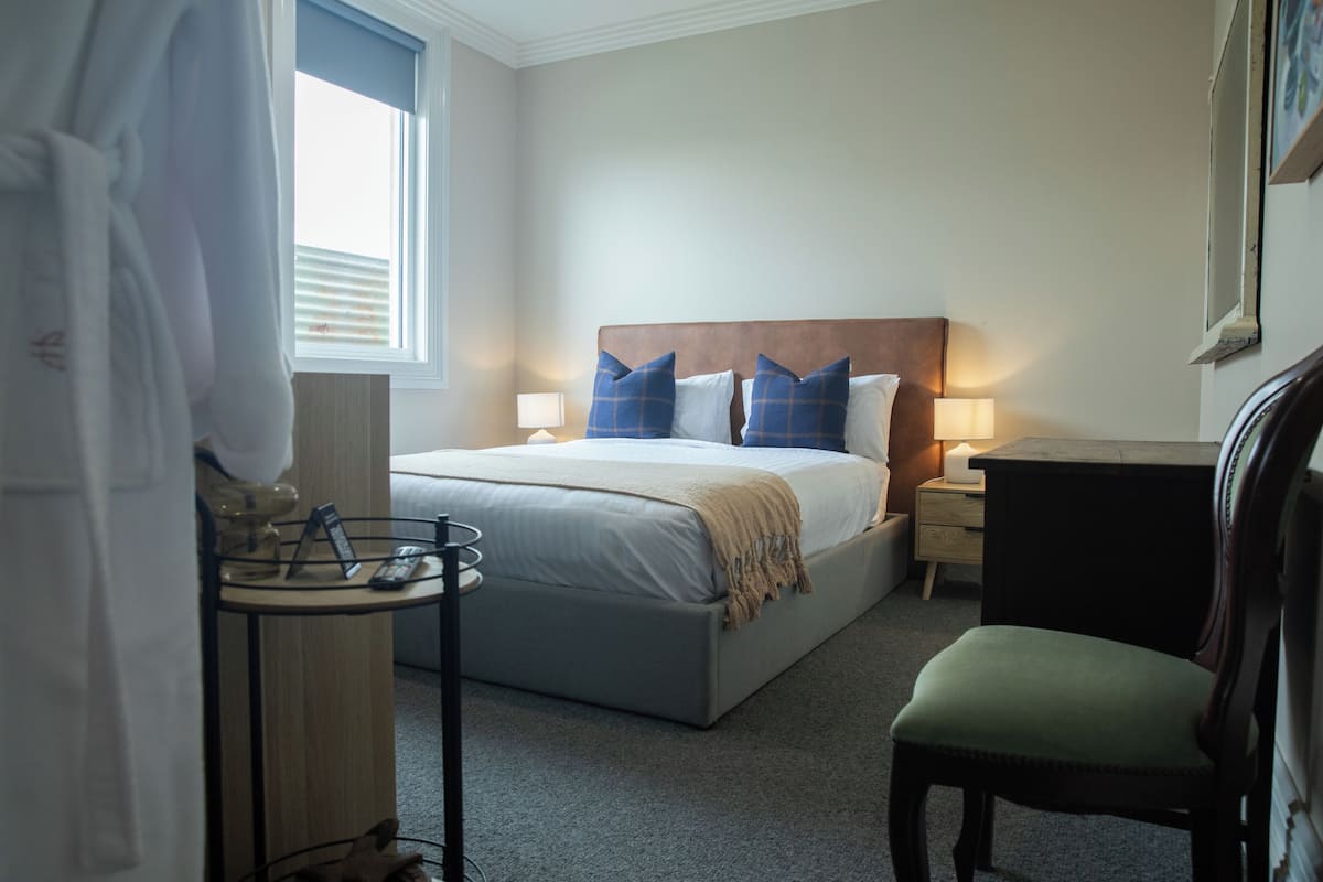 Quarto de hotel com cama, luminárias de cabeceira, uma cadeira, uma mesinha com abajur e uma janela com persianas.