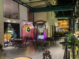Um espaço de refeições interno com cadeiras e mesas de metal decoradas, um letreiro de neon e vista para a rua.