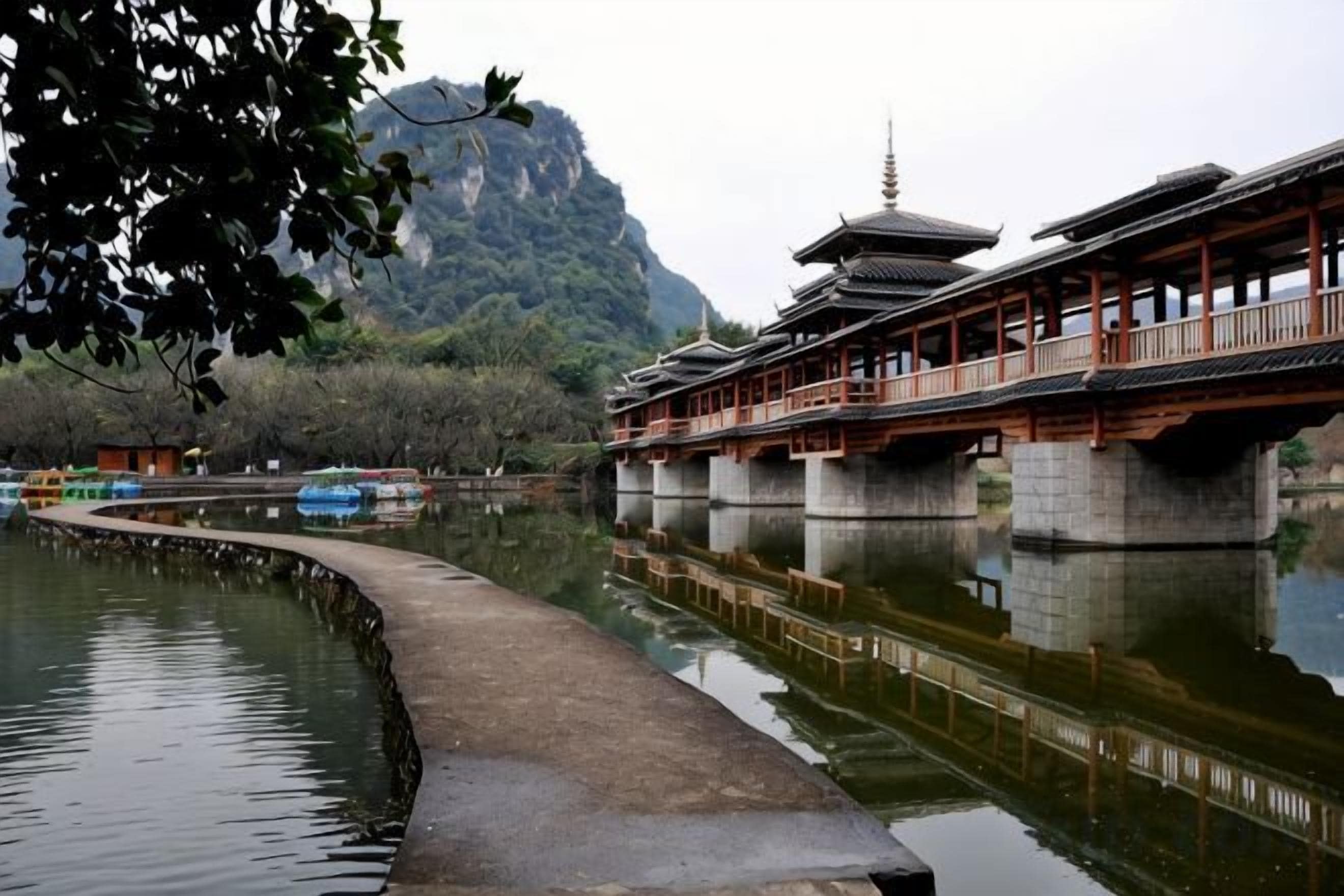 Um edifício tradicional refletido na água, um caminho de pedra e um cenário montanhoso ao fundo.