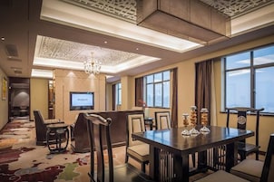 Quarto de hotel com uma grande mesa de jantar, várias cadeiras, um candelabro, uma televisão e vista para a cidade.