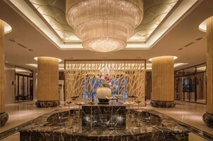 Um hall amplo com balcão de recepção em mármore, um lustre imponente e uma parede decorativa com elementos verticais.