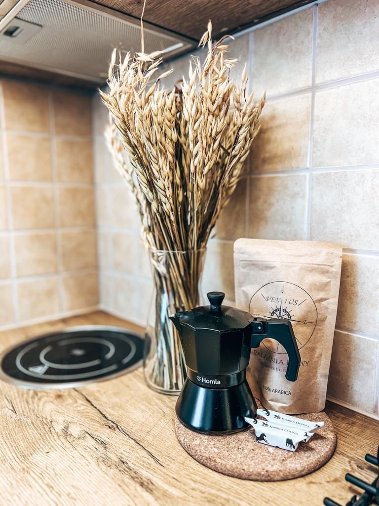 Uma bancada de cozinha com uma cafeteira, um saco de grãos de café e um vaso com espigas de trigo.