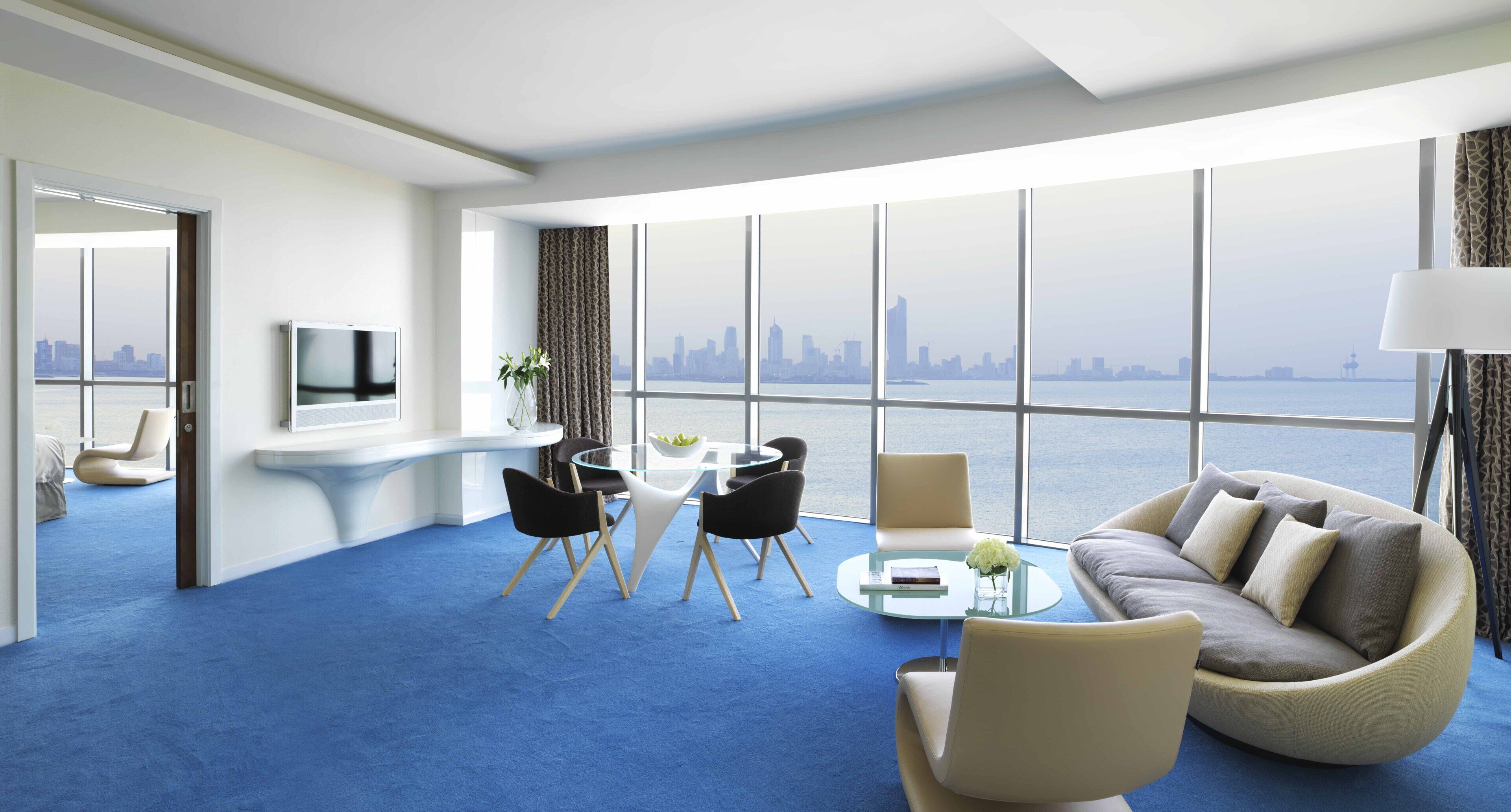 Quarto de hotel moderno com uma grande janela que oferece vista para o horizonte da cidade, uma mesa de jantar redonda com cadeiras, um sofá e uma TV fixada na parede.