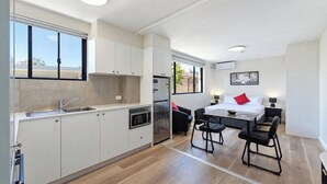 Cozinha moderna com micro-ondas embutido, geladeira e pia. Possui um espaço de refeições com mesa e cadeiras, e uma cama com travesseiro vermelho.
