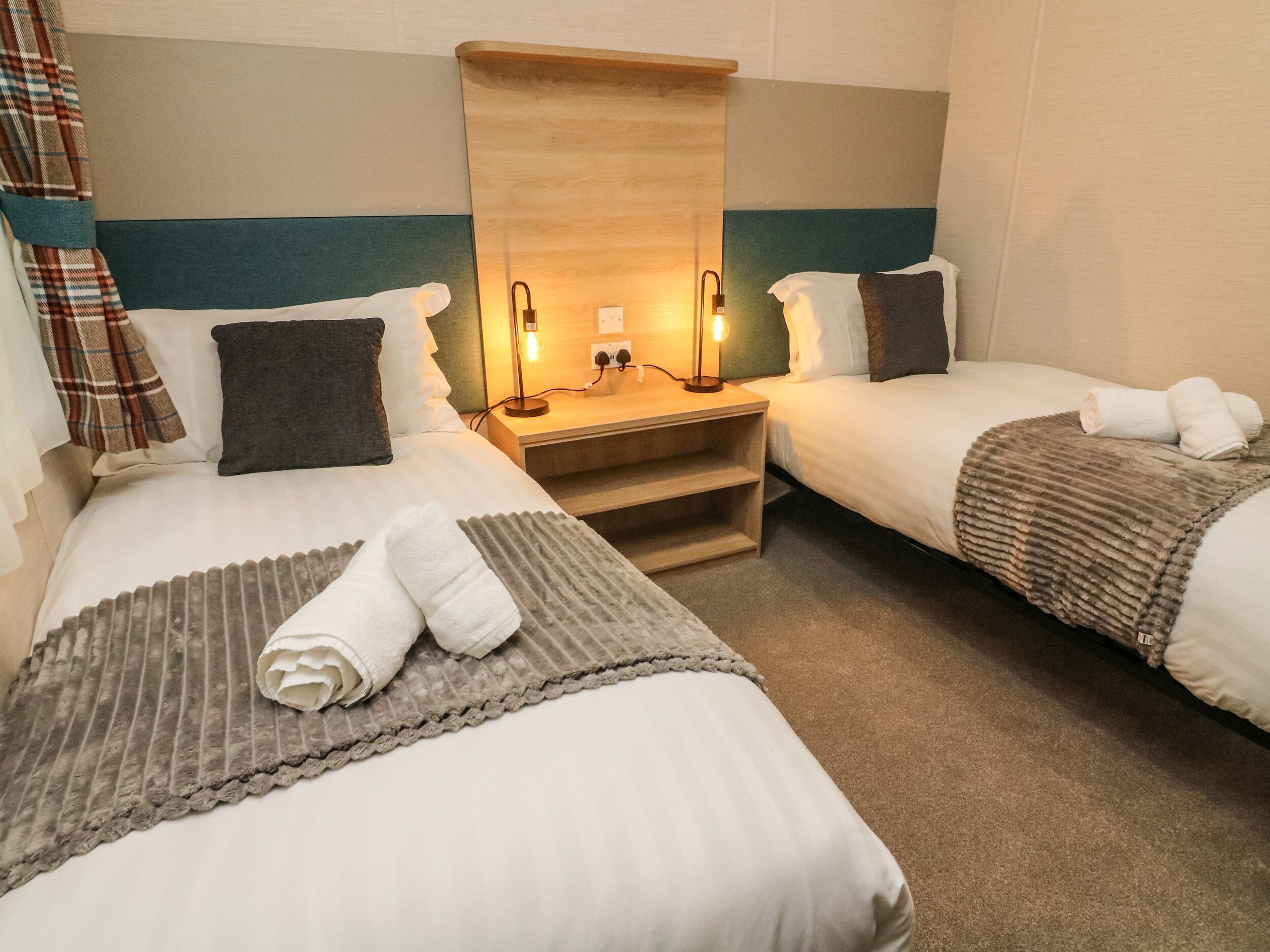 Quarto de hotel com duas camas de solteiro, cada uma com travesseiro, mesa de cabeceira com luminária e manta.