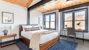 Quarto com cama de madeira, escrivaninha e uma janela grande com vista para uma paisagem seca.