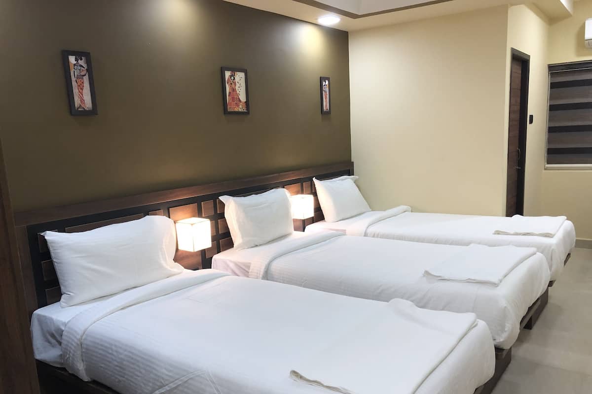 Quarto de hotel com três camas de solteiro, cada uma com roupa de cama branca e cabeceira de madeira. Há luminárias fixadas na parede em ambos os lados das camas.