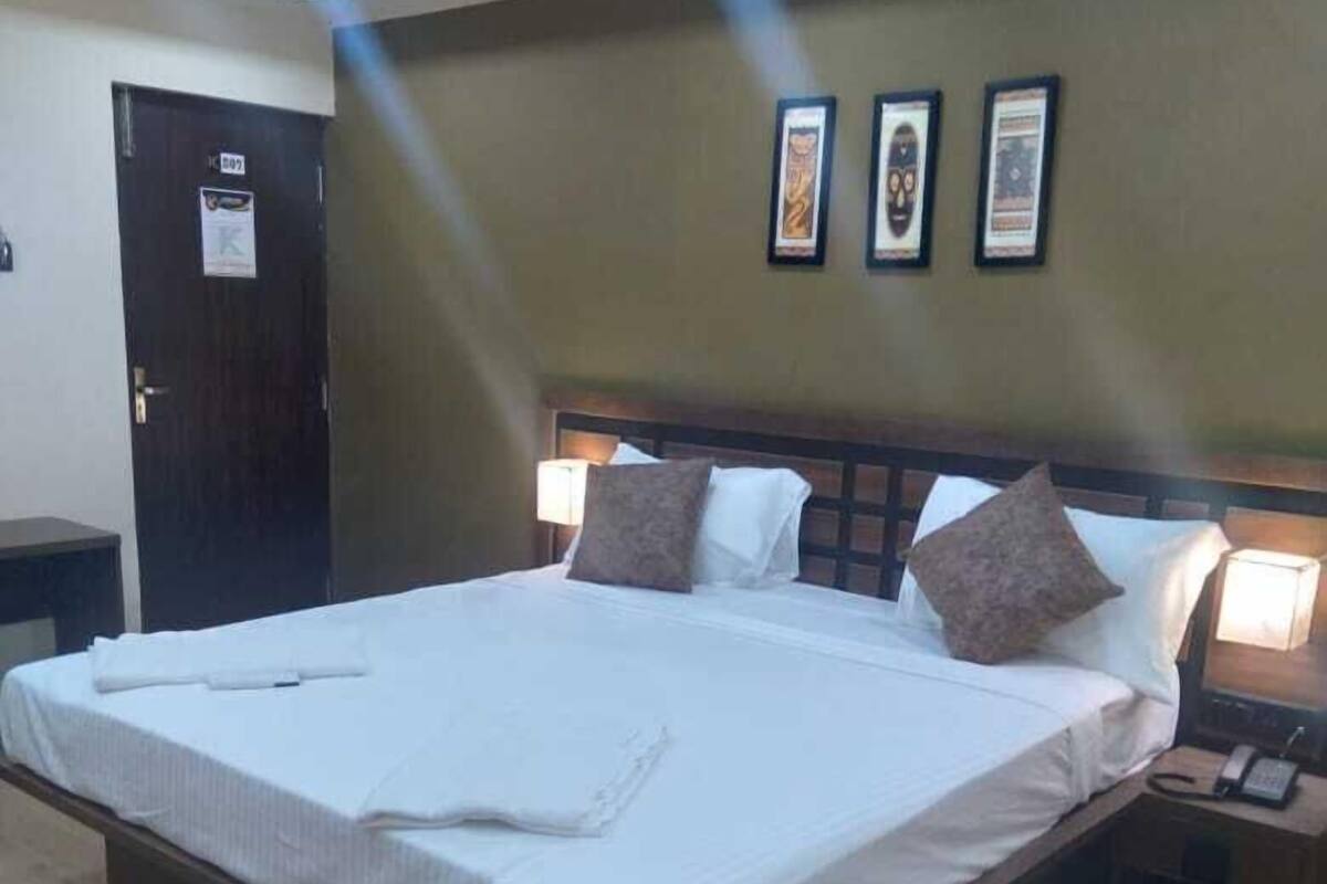 Uma cama grande com estrutura de madeira, duas mesas de cabeceira com abajures, e uma porta com a placa 'K'.