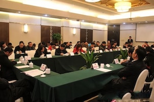 Sala de conferências com mesas compridas, placas de identificação e participantes sentados.