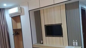 Quarto de hotel com televisão presa a uma parede de painel de madeira, um pequeno armário com produtos de higiene pessoal e um guarda-roupa com cabideiro.