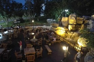 Uma área de estar ao ar livre, com mesas e bancos, rodeada por uma formação rochosa e uma cascata.