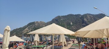 Uma praia com fileiras de espreguiçadeiras e guarda-sóis, um cenário montanhoso ao fundo e céu claro.