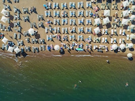 Vista aérea de uma praia com fileiras de espreguiçadeiras e guarda-sóis, com a vastidão do oceano visível e algumas pessoas na água.