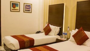Quarto de hotel com duas camas de solteiro, cada uma com colcha e almofadas vermelhas e brancas. Há quadros nas paredes, uma mesa de cabeceira com abajur e uma garrafa de água na mesa.