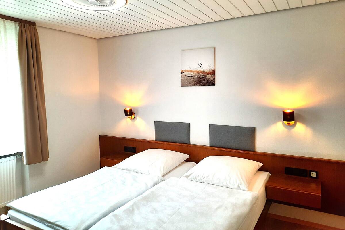 Quarto de hotel com duas camas de solteiro, cabeceira com prateleira embutida, ventilador de teto e duas mesas de cabeceira fixadas na parede com luminárias.