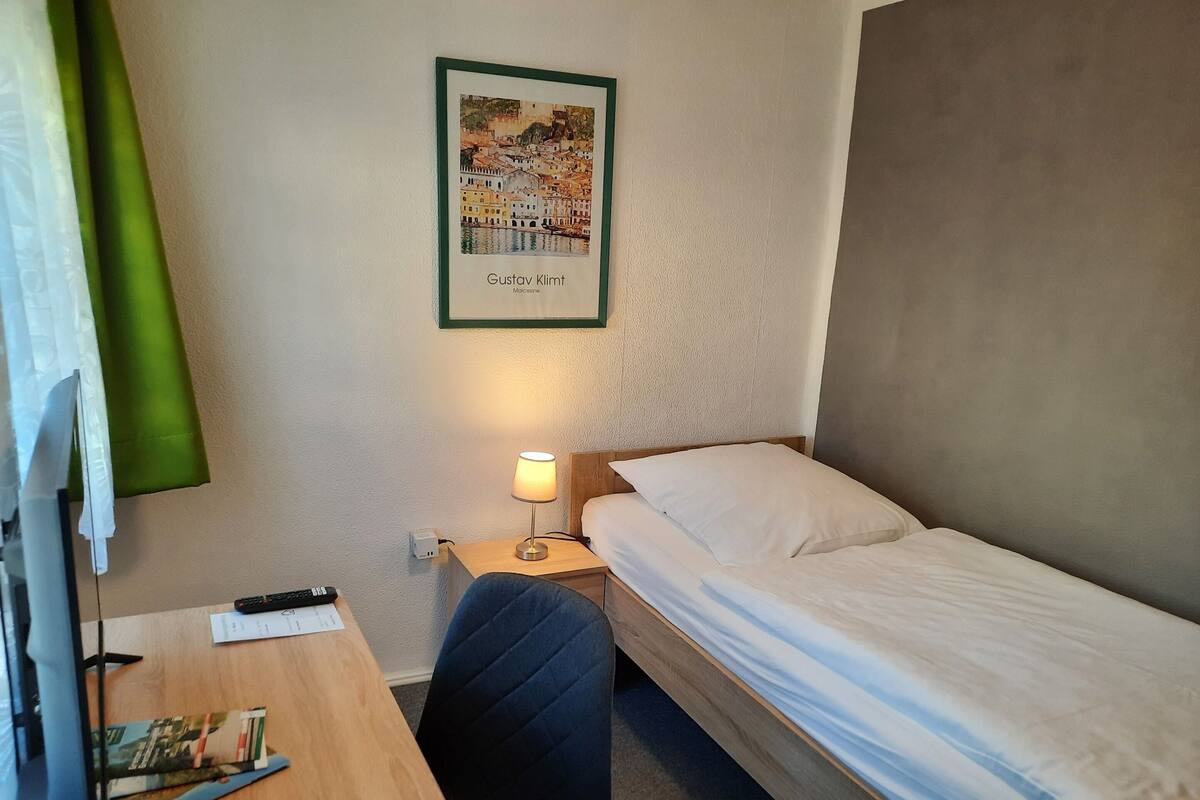 Quarto de hotel com uma cama, uma escrivaninha com abajur, um quadro na parede e uma cortina verde.