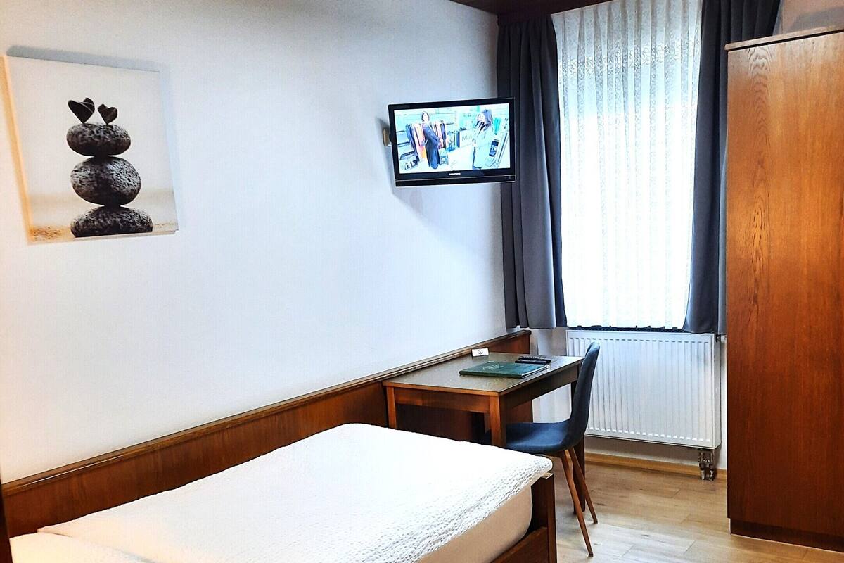 Quarto com cama, escrivaninha, cadeira, televisão e janela com cortinas.