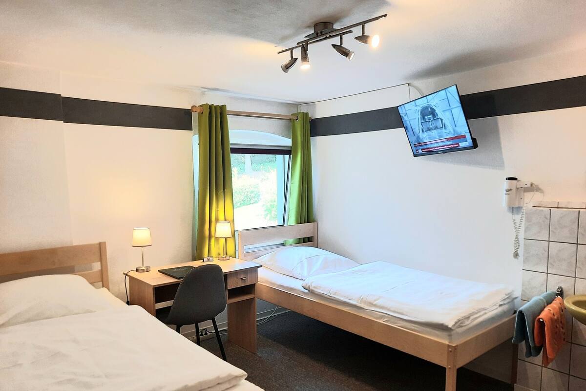 Quarto de hotel com duas camas, uma escrivaninha com cadeira, televisão na parede e janela com cortinas.