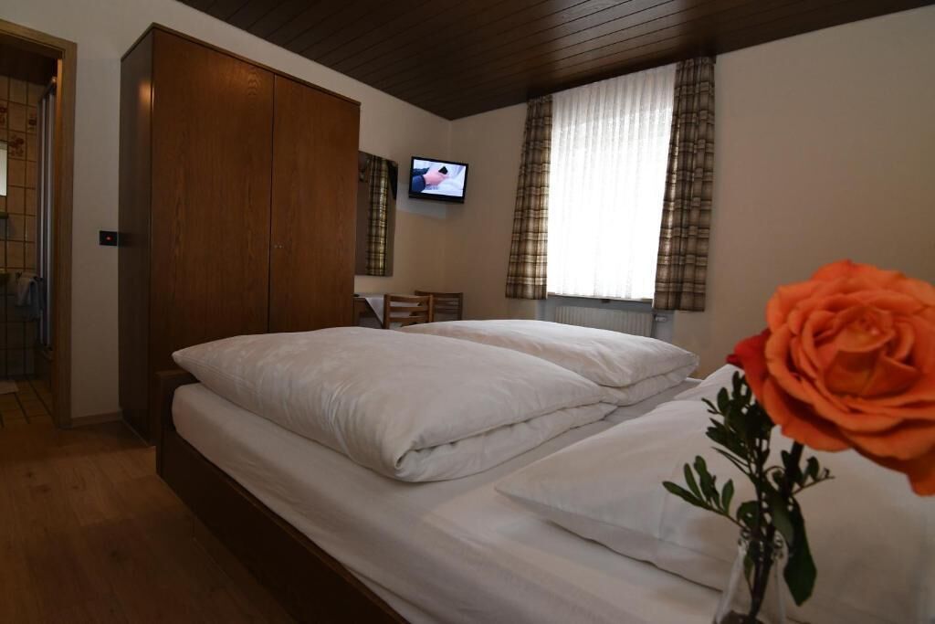Quarto com guarda-roupa de madeira, cama com roupa de cama branca, televisão na parede e janela com cortinas.
