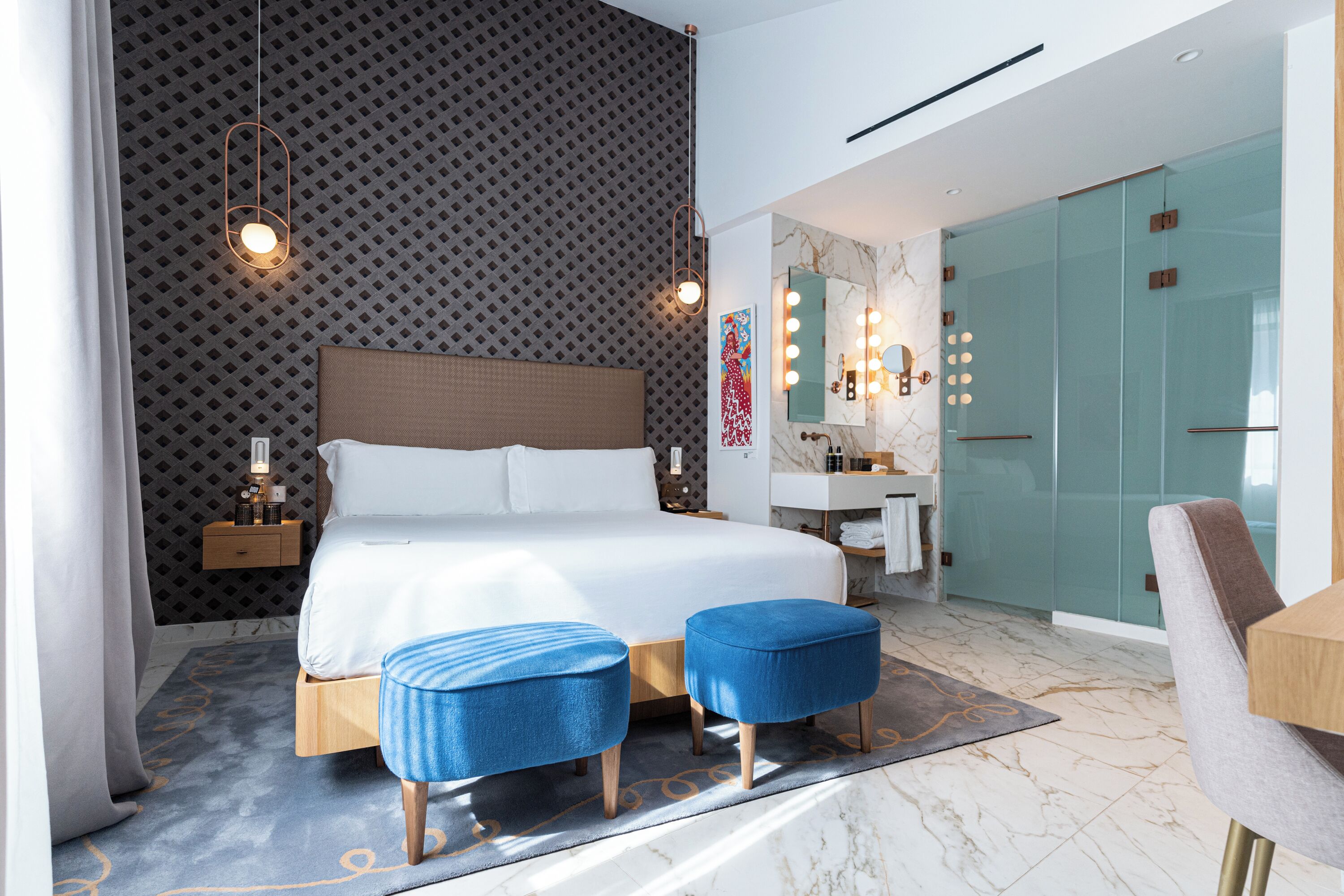 Quarto de hotel moderno com uma cama grande, dois pufes azuis, uma toucador com espelho e iluminação, e um closet espaçoso.