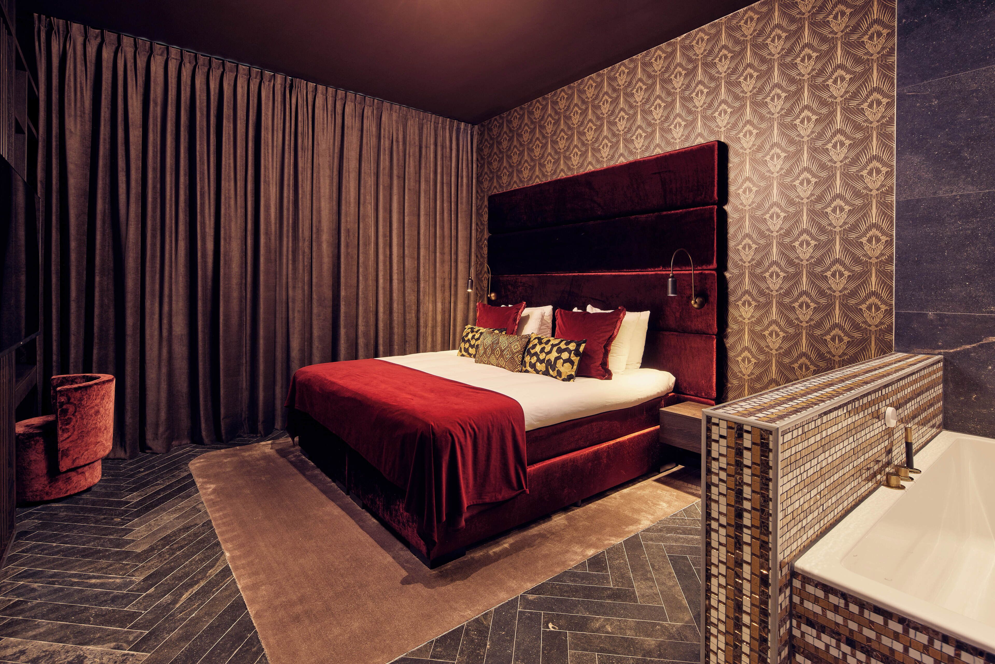 Quarto com cama grande, cabeceira vermelha, parede estampada e banheiro com área de chuveiro revestida em azulejos.