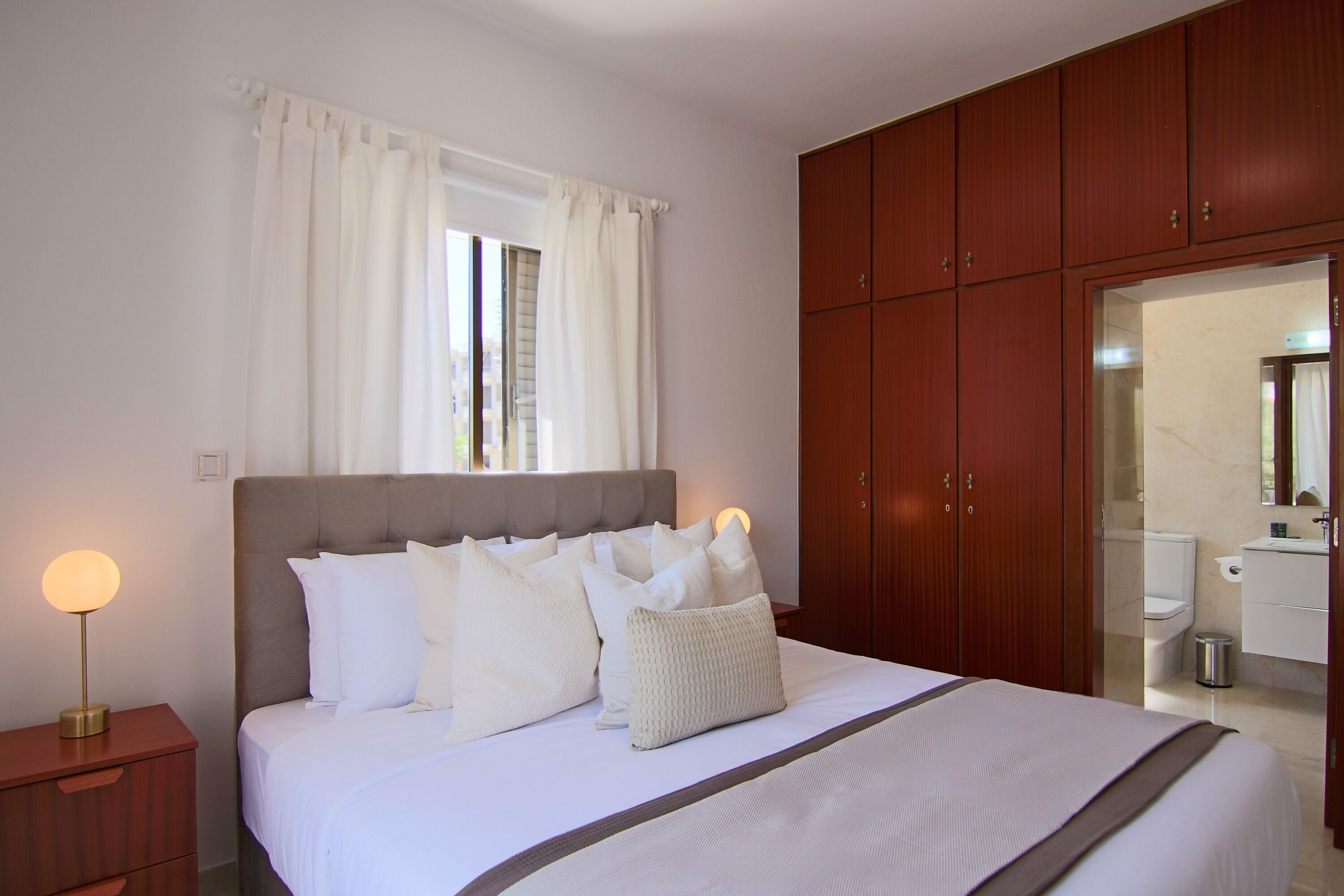 Uma cama bem arrumada com roupa de cama branca e várias almofadas, uma mesa de cabeceira com abajur e um guarda-roupa de madeira.