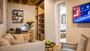 Uma sala de estar com sofá, poltrona e uma mesinha com uma bandeja de maçãs. Há uma estante de livros, uma televisão fixada na parede e uma porta que dá para uma varanda.