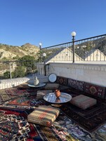 Terraço no telhado com vista para uma paisagem montanhosa, com guarda-corpo de ferro forjado, vários tapetes coloridos e um pote de barro tradicional.