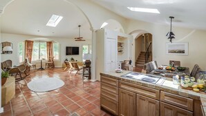 Uma cozinha ampla com ilha central, uma sala de jantar com mobiliário de vime e uma escada que dá para um andar superior.