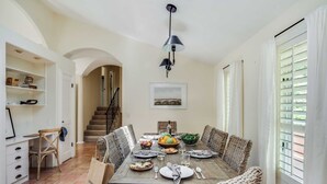 Uma sala de jantar com uma mesa de madeira posta para seis pessoas, cercada por cadeiras de vime, um lustre e uma escadaria que leva a outro andar.