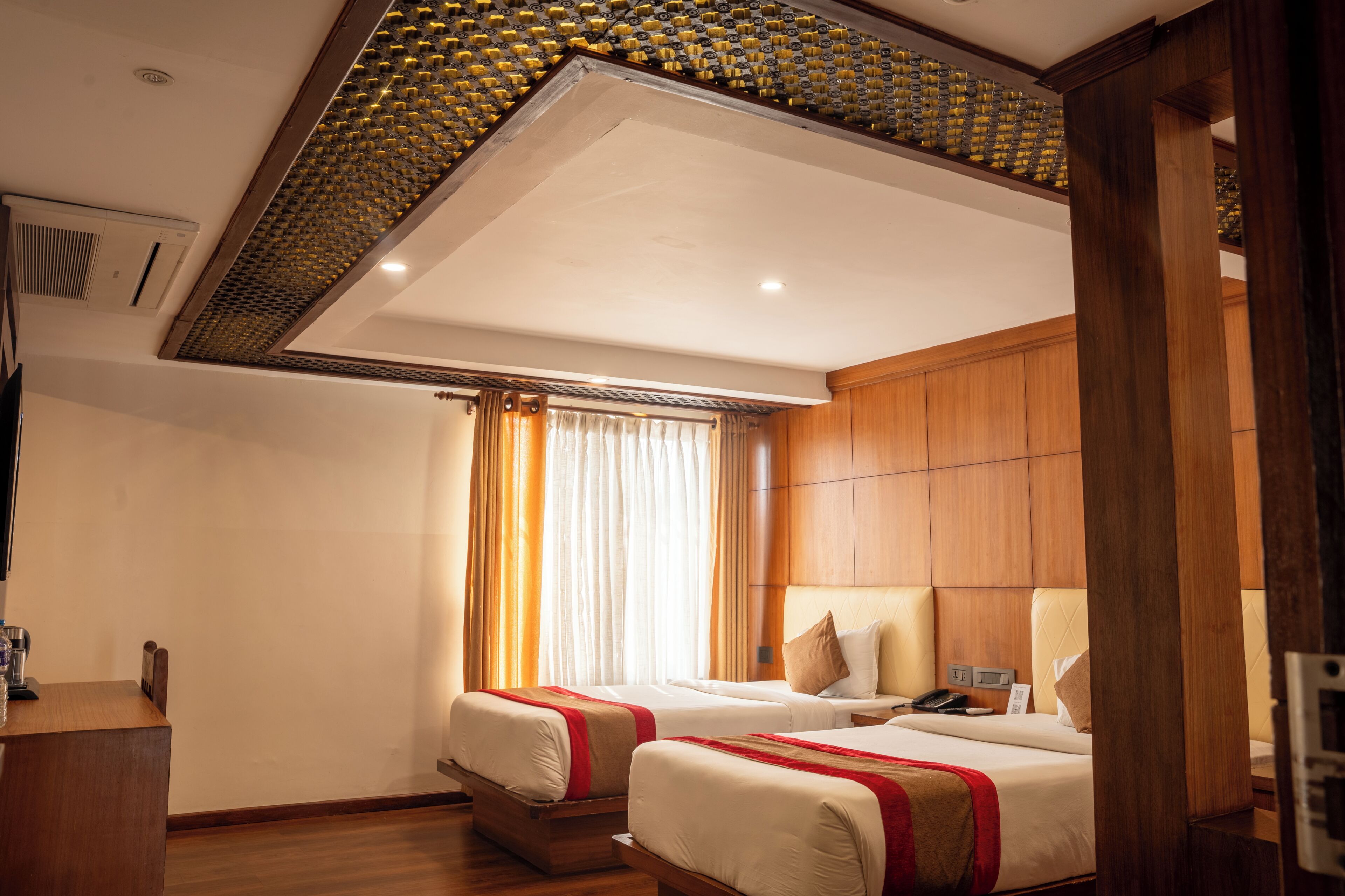 Quarto de hotel com duas camas, painéis de madeira, uma janela grande com cortinas e teto com padrão decorativo.