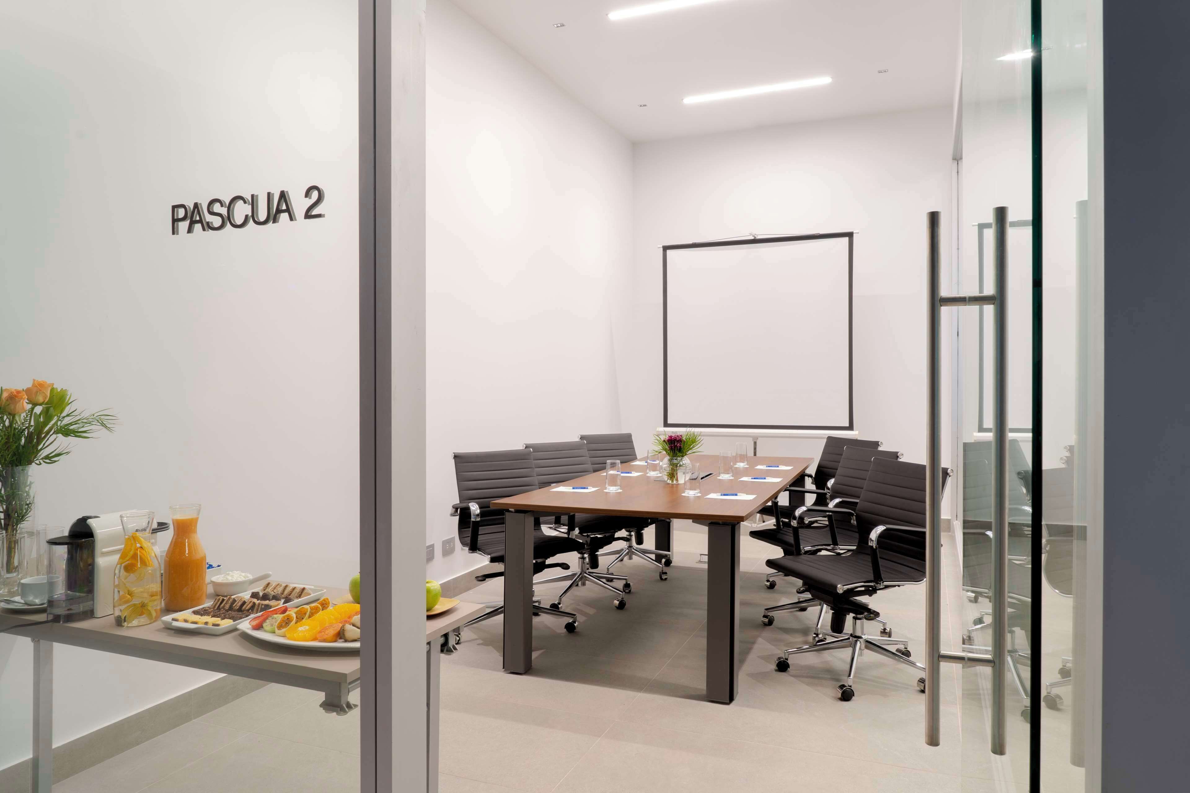 Sala de conferências com uma mesa grande, cadeiras, tela de projetor e divisória de vidro.