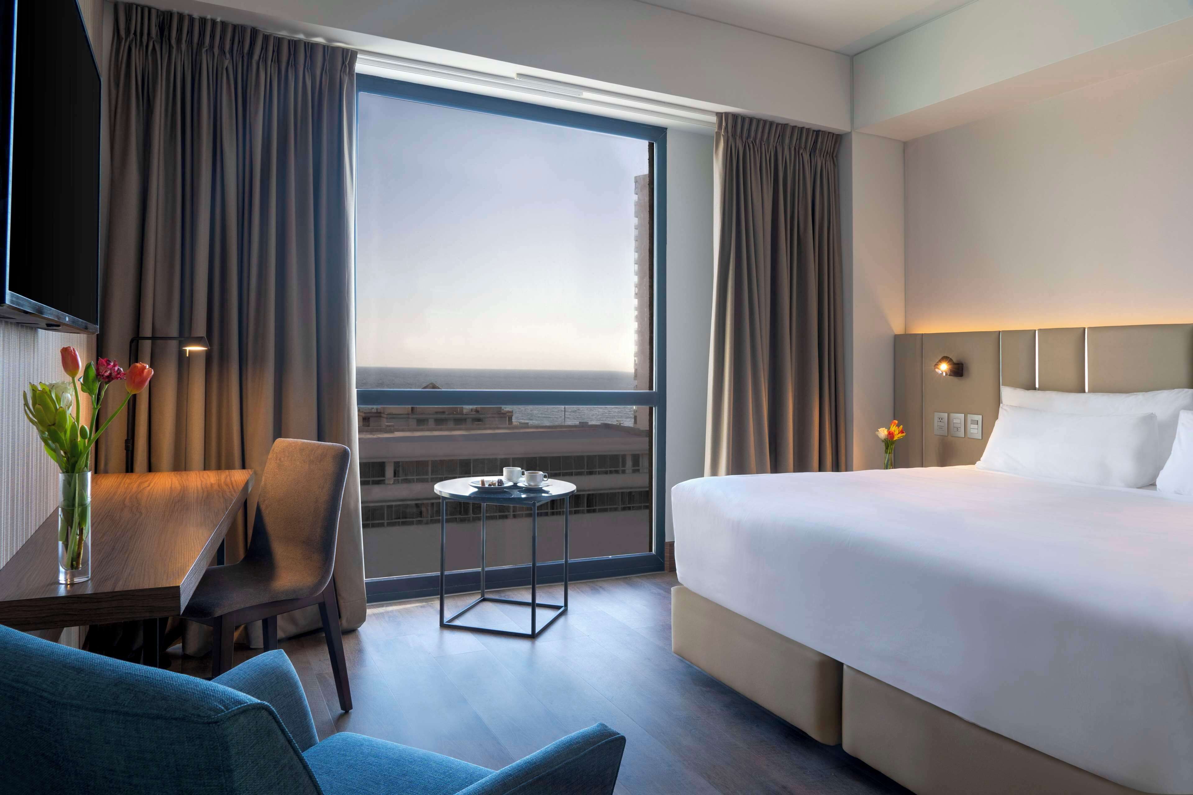Quarto de hotel com uma cama grande, uma mesa com cadeira, uma mesinha e vista da cidade através de grandes janelas.