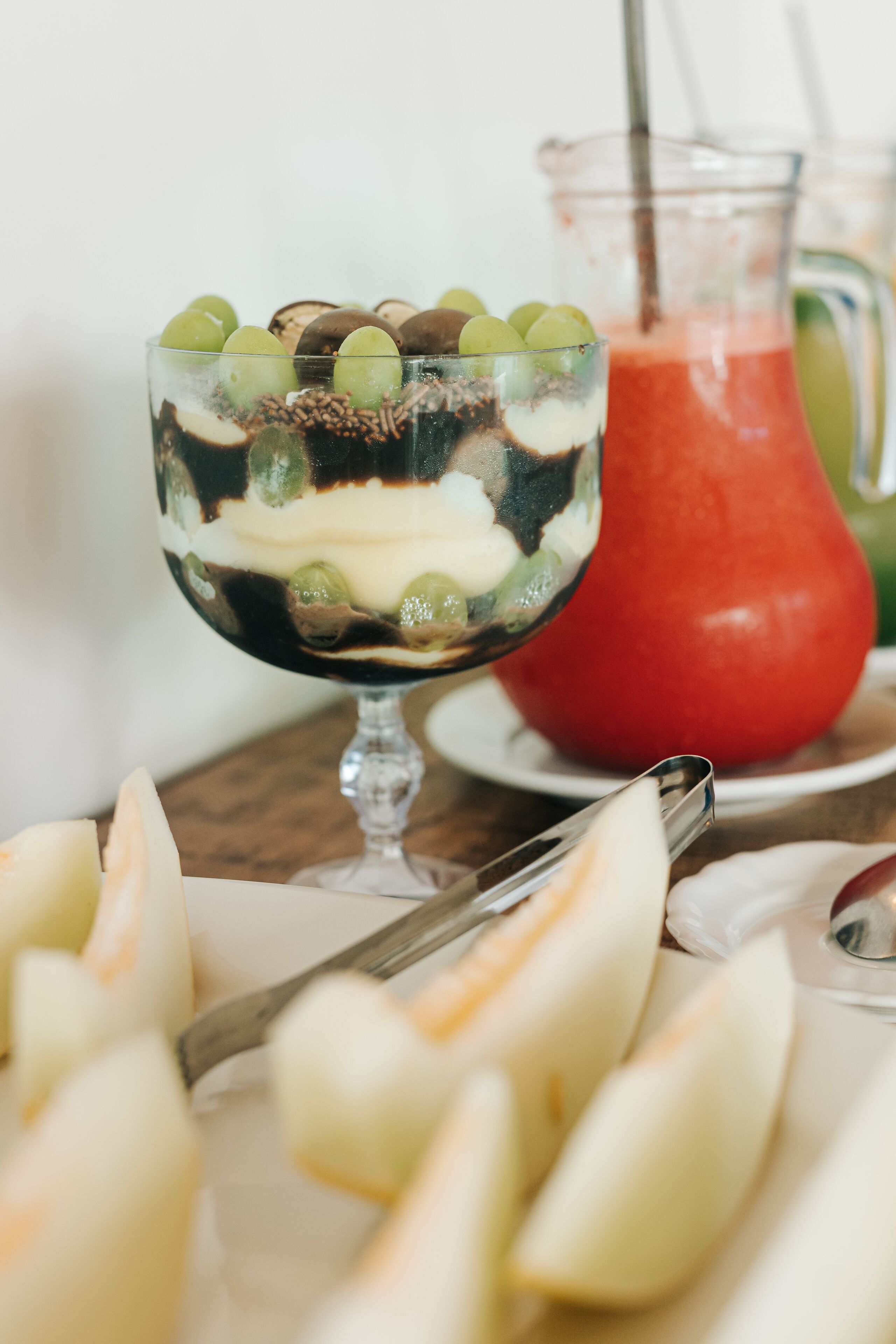 Um copo de sobremesa com camadas de bolo, fruta e creme, uma jarra de bebida vermelha e uma colher sobre a mesa.