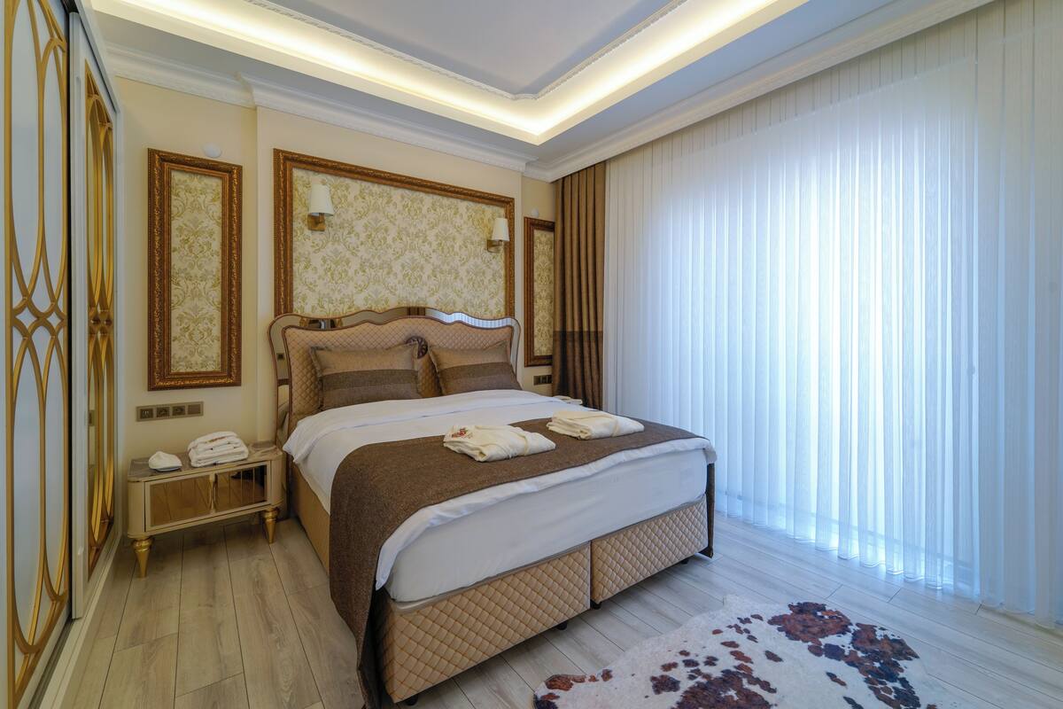 Um quarto espaçoso com uma cama grande, mesa de cabeceira, espelho e janela com cortinas finas.