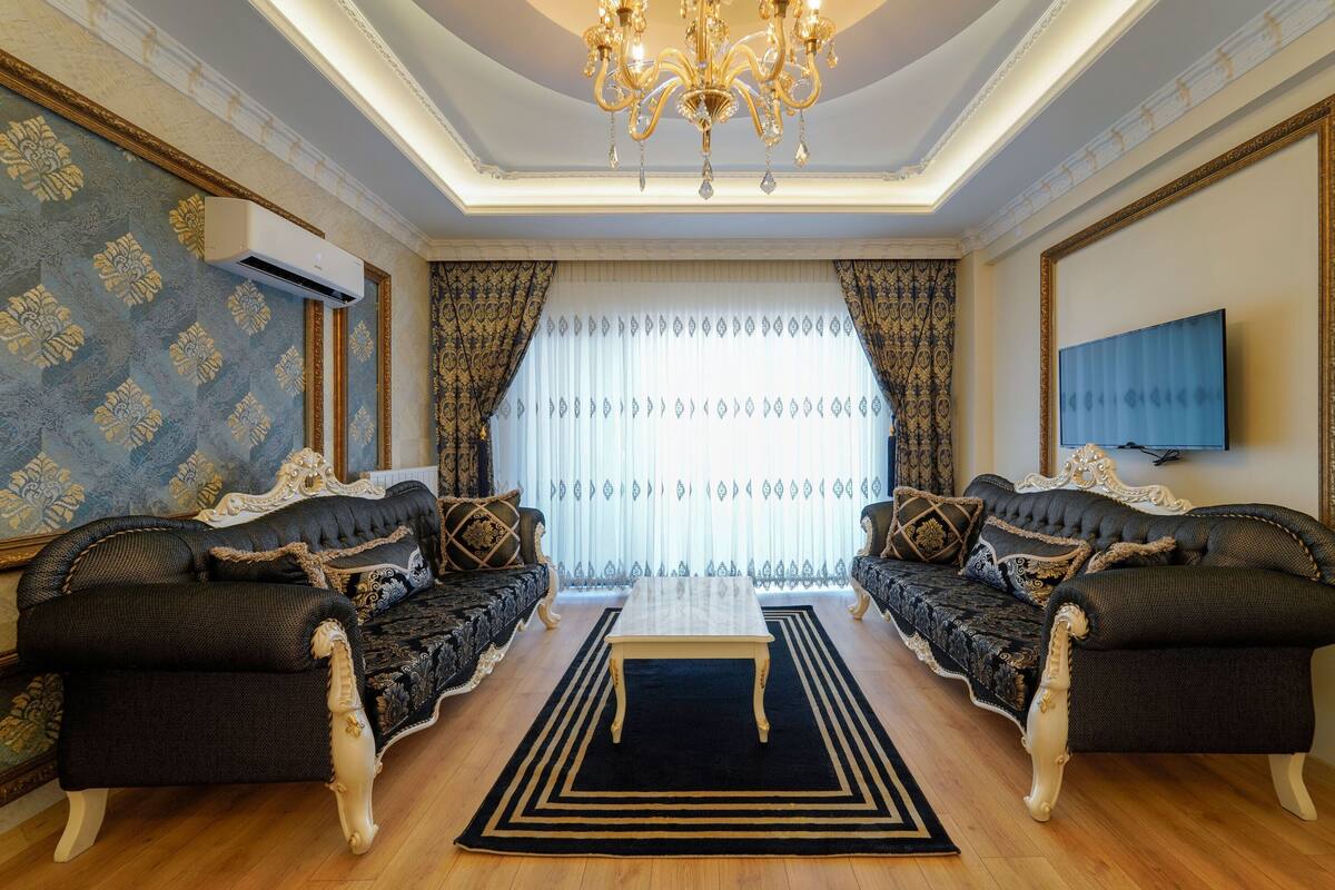 Uma sala de estar luxuosa com sofás ricamente decorados, um candelabro e uma grande janela com cortinas estampadas.