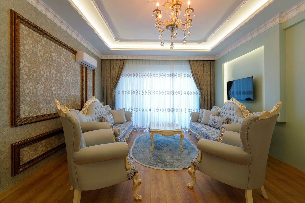 Uma sala de estar ampla com móveis elaborados, um candelabro e uma grande janela com cortinas estampadas.