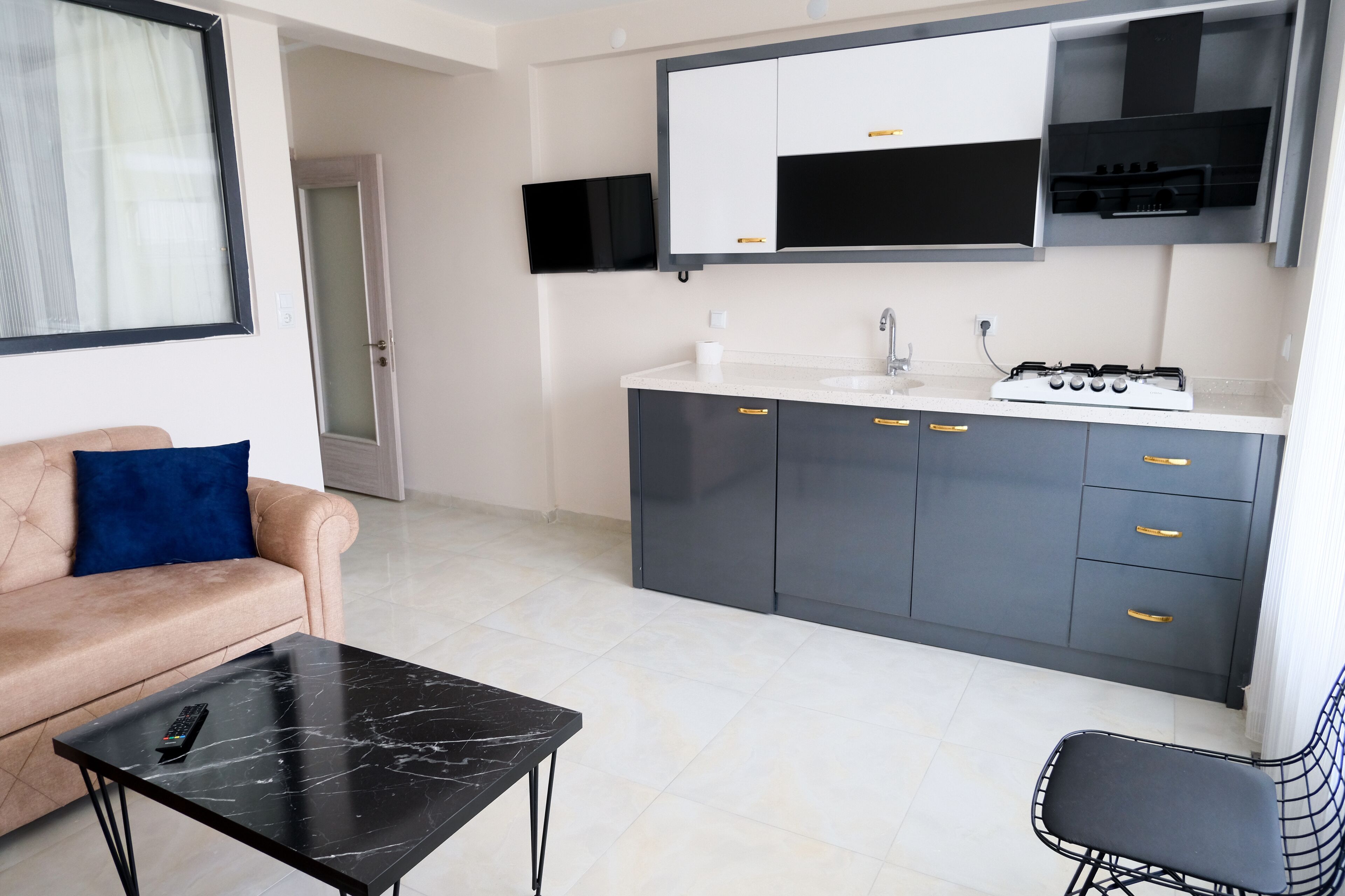Uma cozinha moderna com armários cinza, bancada branca e fogão preto. Há um sofá bege com almofada azul e uma mesa de centro preta com um controle remoto.