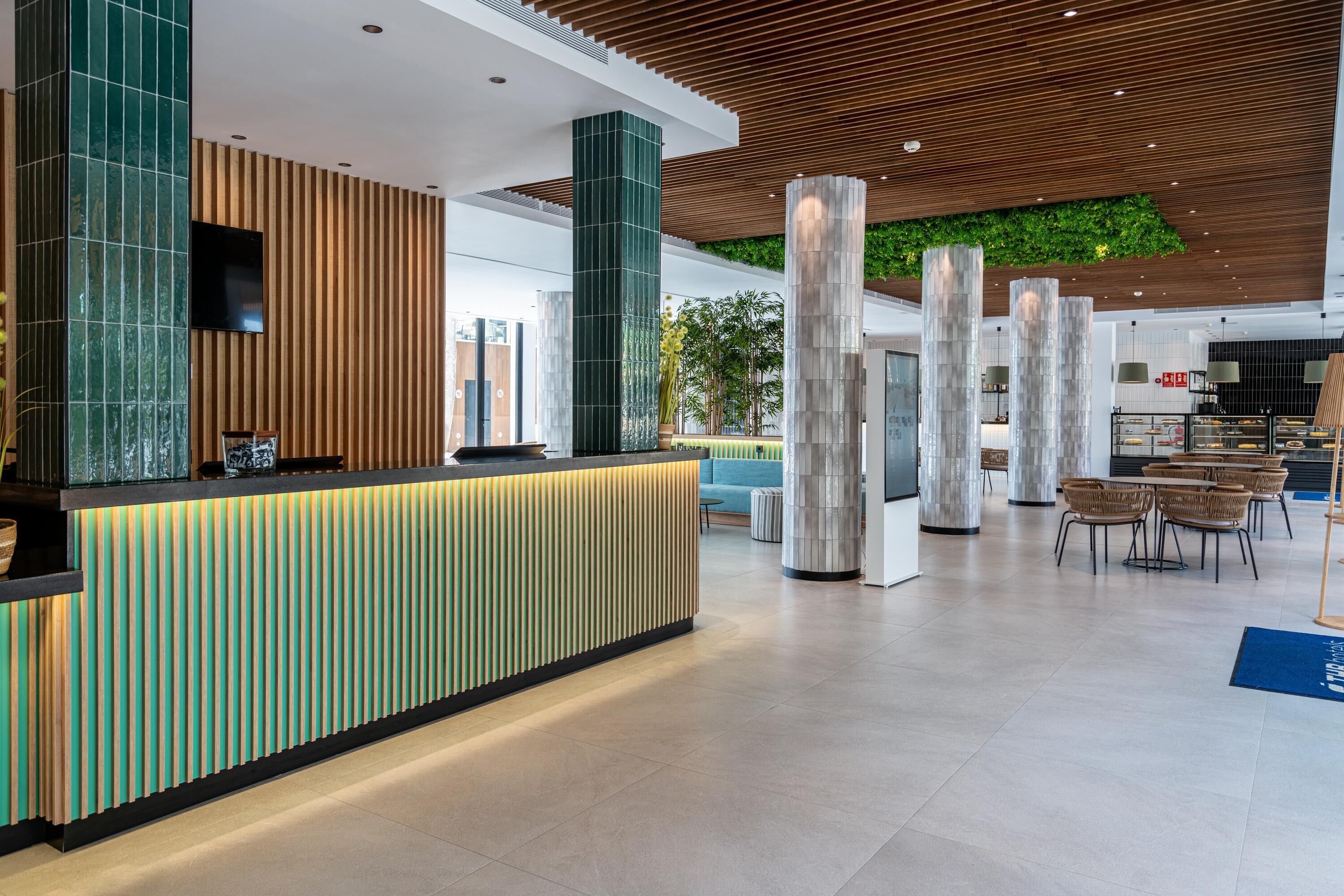 Uma área de recepção moderna com um balcão de atendimento em padrão verde e amarelo, painéis de madeira vertical e uma parede verde com planta.