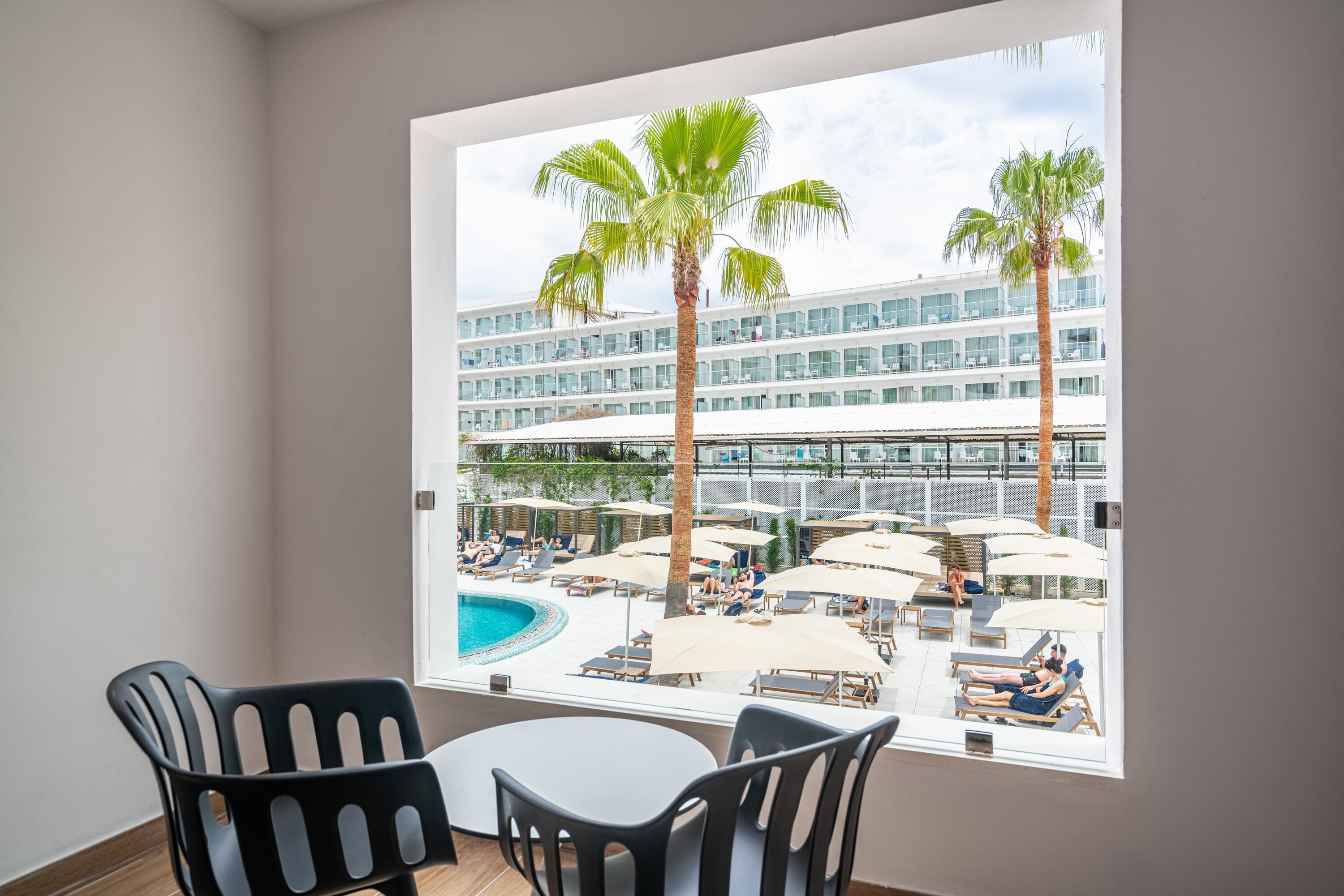 Quarto com uma grande janela com vista para uma área de piscina com espreguiçadeiras e palmeiras.