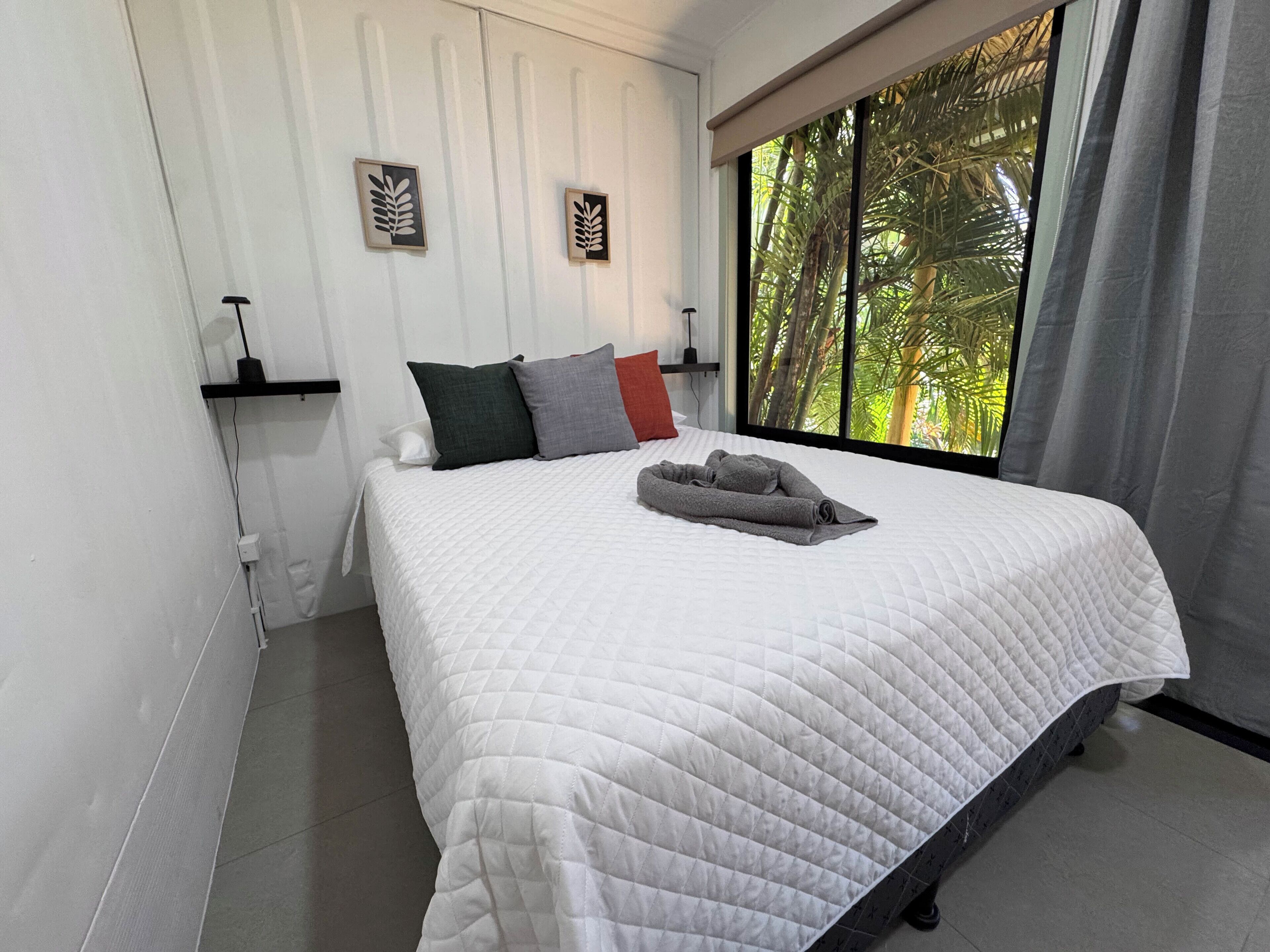 Cama bem arrumada com colcha branca, dois travesseiros e uma toalha enrolada. Dois quadros na parede e uma janela com vista para o verde.