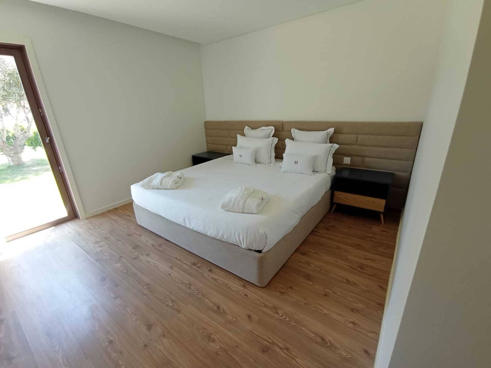 Quarto moderno com cama grande, piso de madeira e vista para o exterior.