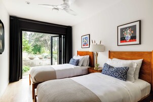 Quarto de hotel com duas camas, ventilador de teto, janela com cortinas e quadros nas paredes.