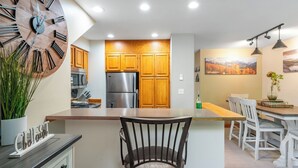 Cozinha moderna com armários de madeira, uma geladeira de aço inoxidável e um espaço de jantar com cadeiras brancas e uma mesa.