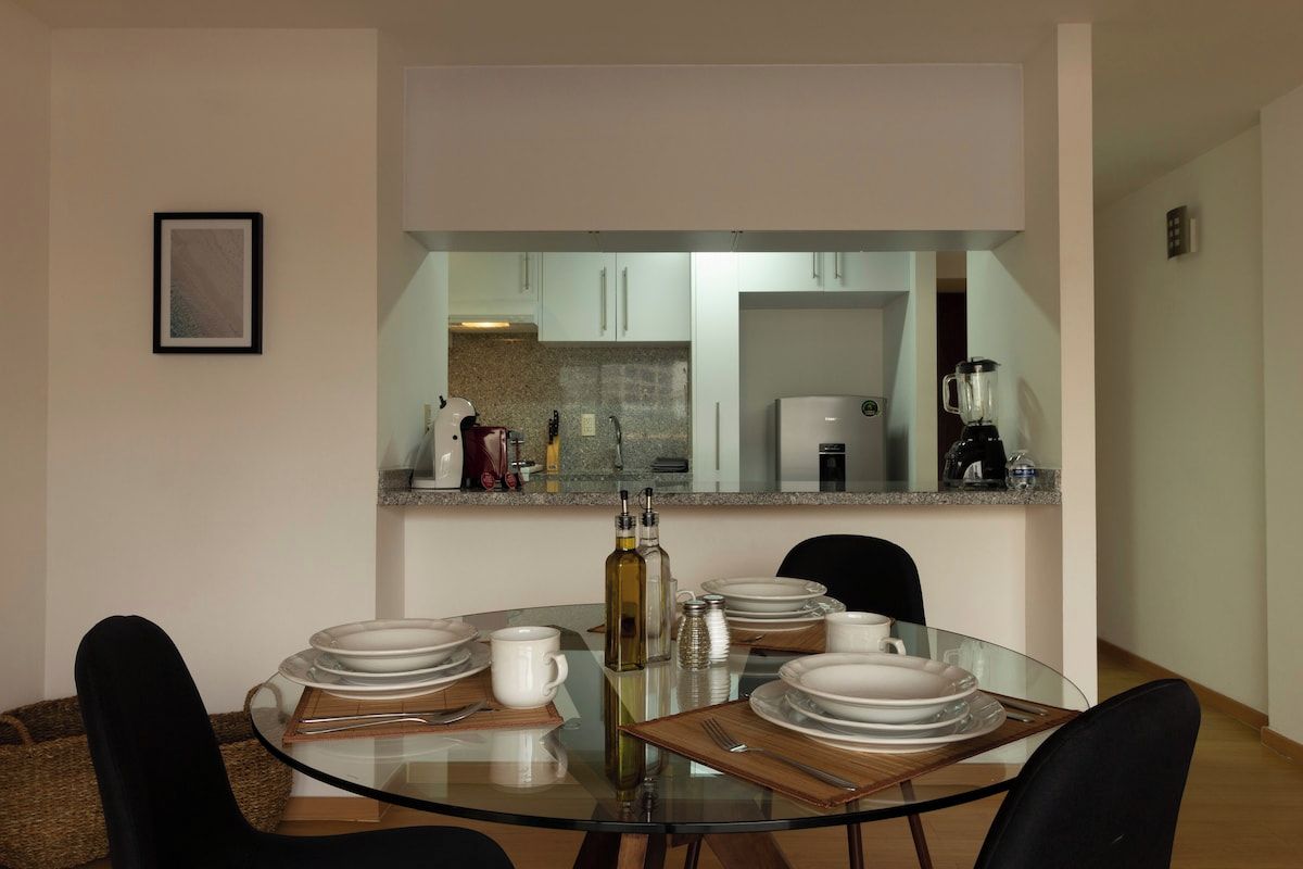 Uma sala de jantar com mesa de vidro, cadeiras pretas e cozinha ao fundo.
