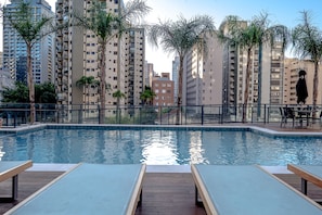 Piscina com espreguiçadeiras de madeira, cercada por edifícios residenciais altos e palmeiras.