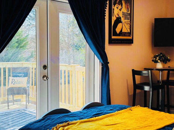 Quarto de hotel com uma cama, uma escrivaninha e uma cadeira. Há uma janela com cortinas, um cartaz na parede e vista para um terraço lá fora.
