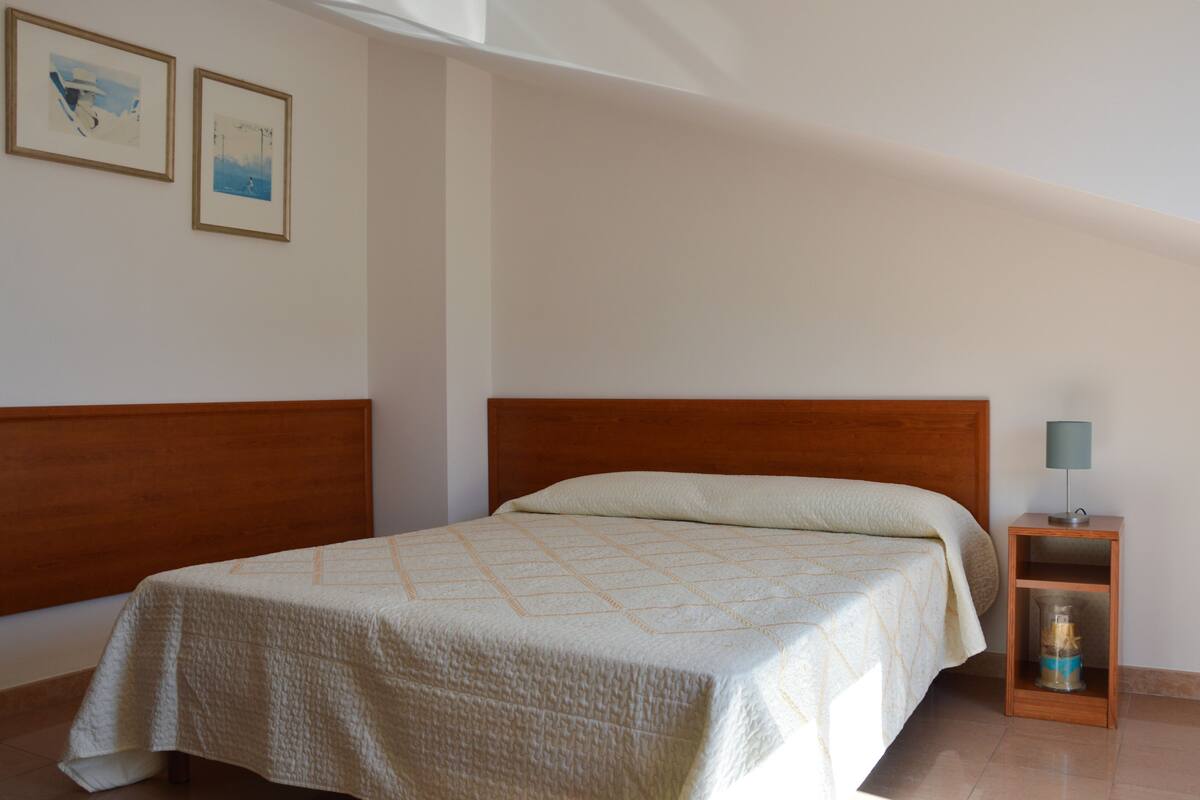Uma cama de solteiro com cabeceira de madeira, uma mesa de cabeceira com abajur e duas imagens emolduradas na parede.